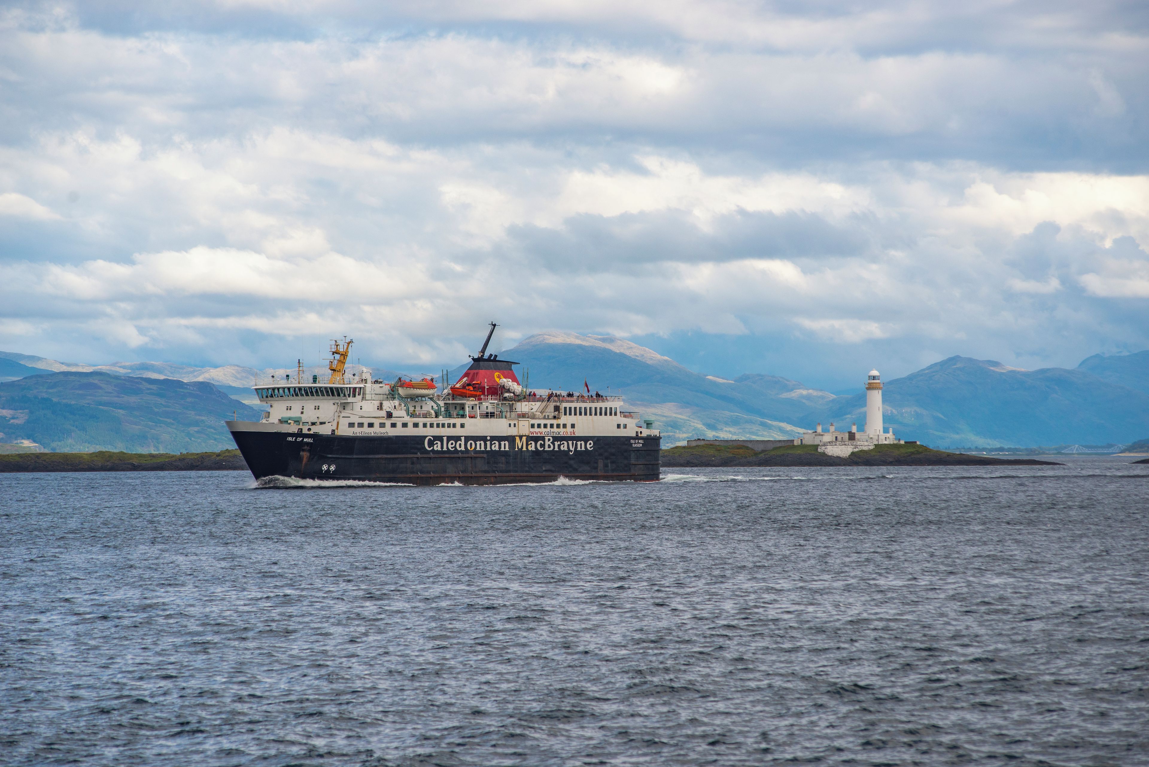 Um grande navio de cruzeiro chamado 'Caledonian MacBrayne' navegando em águas com paisagem montanhosa e um farol ao fundo.