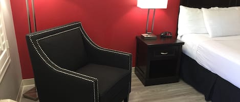 Quarto de hotel com poltrona estofada preta, mesa de cabeceira de madeira com abajur, cama com roupa de cama branca e parede vermelha.