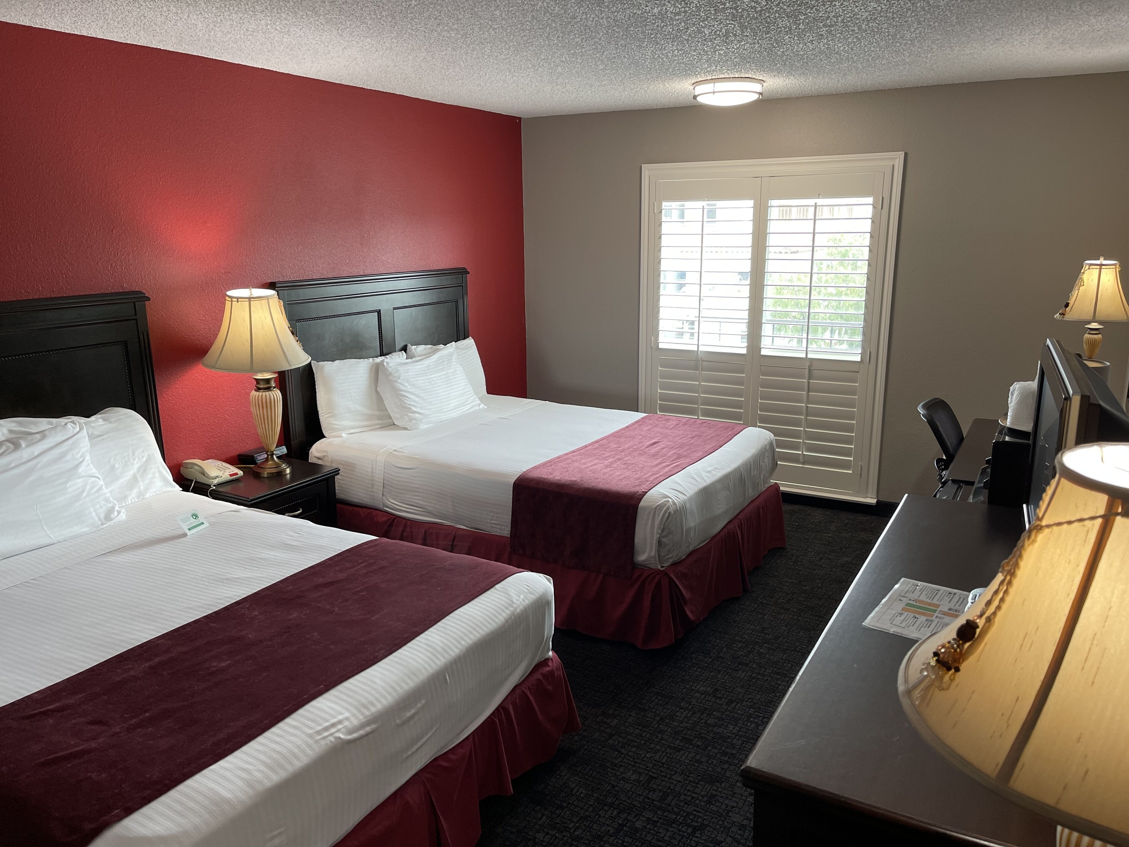Quarto de hotel com duas camas, uma escrivaninha, uma cadeira, um abajur e uma janela com persianas.