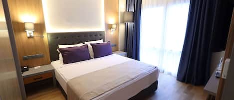 Quarto de hotel com uma cama grande, uma mesa de cabeceira, uma luminária na parede e uma janela com cortinas.