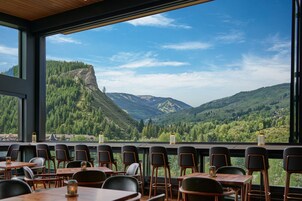 Um restaurante com mesas e cadeiras, grandes janelas com vista para uma paisagem montanhosa.