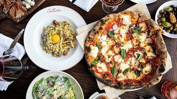 Uma mesa com diversos pratos, incluindo massa, pizza e uma salada, acompanhada de copos de uma bebida escura.