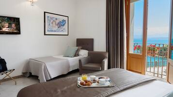 Quarto de hotel com cama, poltrona, mesa pequena com café da manhã, pintura na parede e vista para o mar.