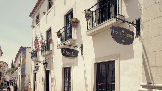 Uma rua de paralelepípedos com um hotel chamado "Hotel Doce Rainha" e um edifício com varandas decoradas com plantas em vasos.