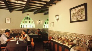 Interior de restaurante com vigas de madeira, paredes azulejadas e um mural. Mesas montadas com pratos e bebidas.