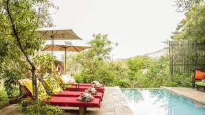 Uma área junto à piscina com cadeiras de descanso vermelhas e um grande guarda-sol, cercada por vegetação e um muro de madeira.