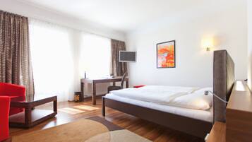 Quarto de hotel com cama, escrivaninha, cadeira, televisão e janela com cortinas.