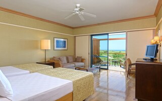 Quarto de hotel com duas camas, um sofá, uma mesa de escritório com um computador e uma varanda com vista.