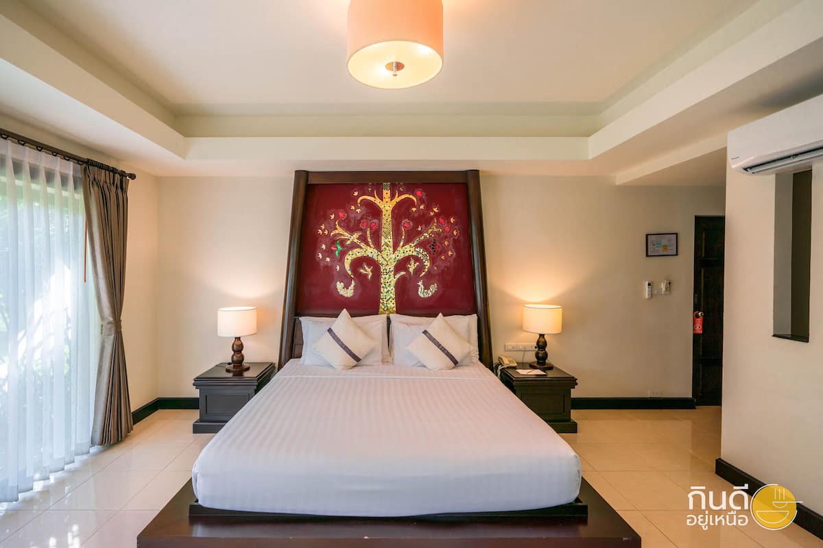 Quarto de hotel com cama grande, duas mesas de cabeceira com luminárias, cabeceira decorativa e vista para o exterior através de cortinas leves.