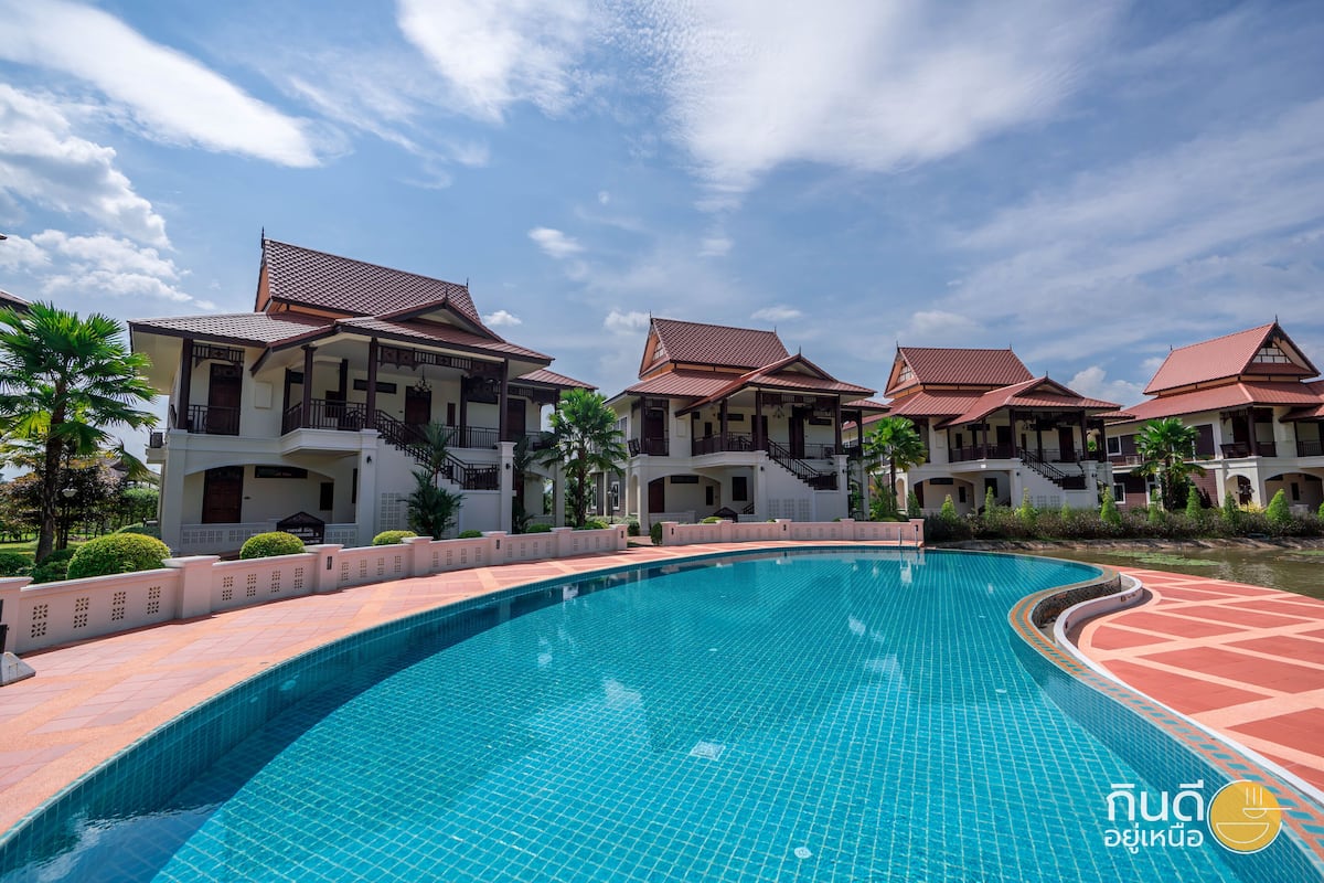 Uma piscina com borda curva, rodeada por um deck revestido e uma fileira de edifícios de estilo tradicional com telhados vermelhos e varandas.