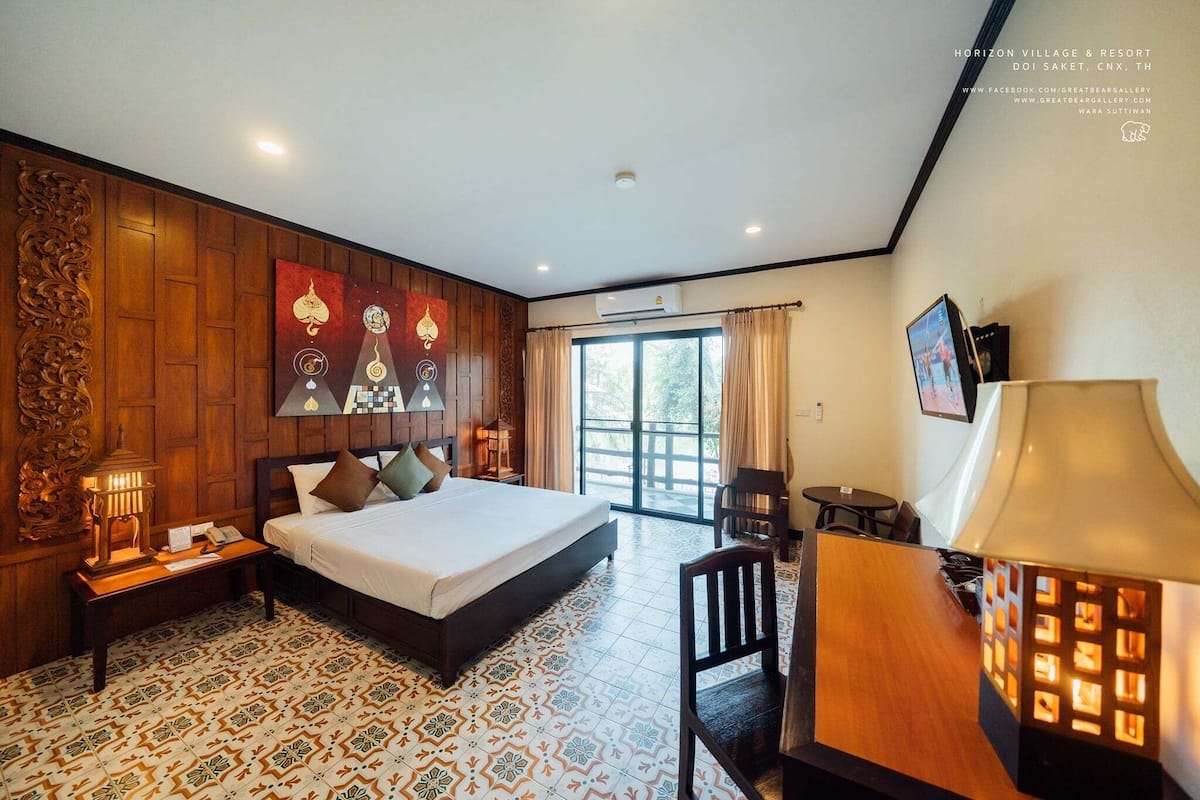 Quarto de hotel com cama, escrivaninha, cadeira, luminária e uma janela grande.