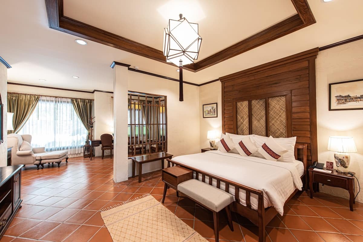 Quarto de hotel com cama grande, cabeceira de madeira, escrivaninha com cadeira, um banquinho, mesa de cabeceira com abajur e vista para o exterior através de uma janela com cortinas.