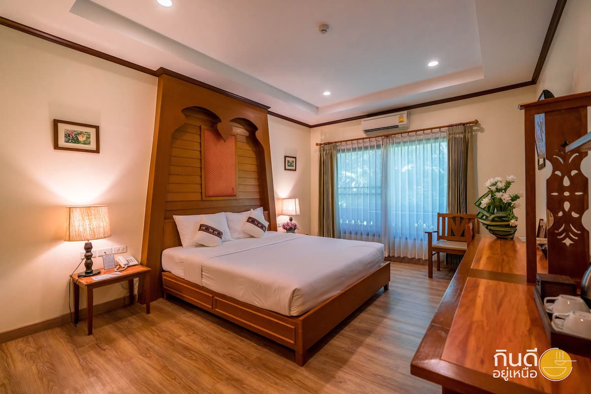 Quarto de hotel com cama de madeira, mesas de cabeceira com abajures, escrivaninha com cadeira e uma janela grande com cortinas.