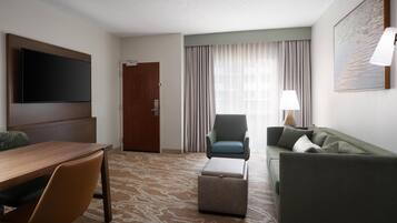 Quarto de hotel com televisão de tela plana, mesa de jantar de madeira com cadeiras, sofá, um pequeno banquinho e uma janela grande com cortinas.