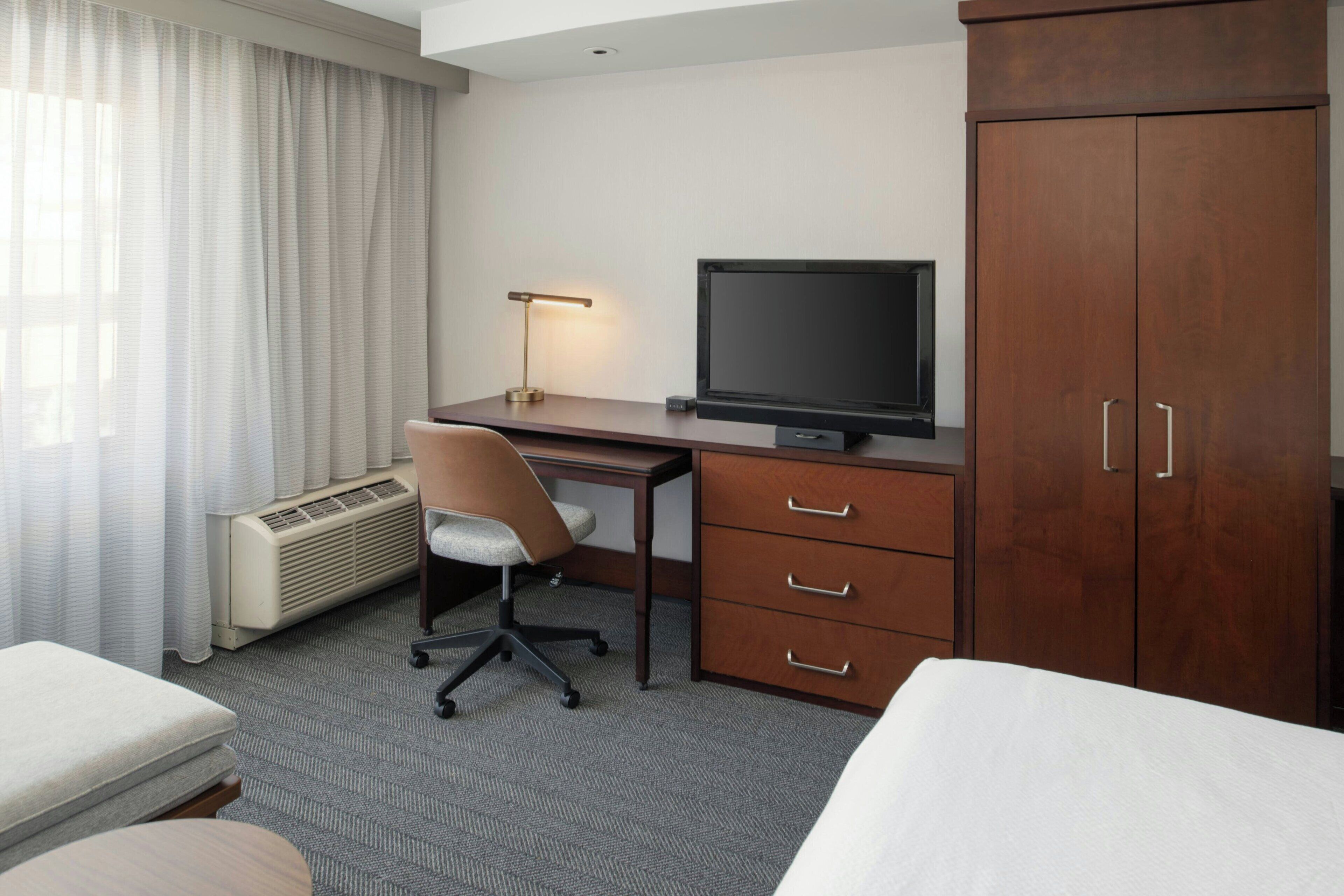 Quarto de hotel com cama, escrivaninha com computador, cadeira, armário e janela com cortinas.