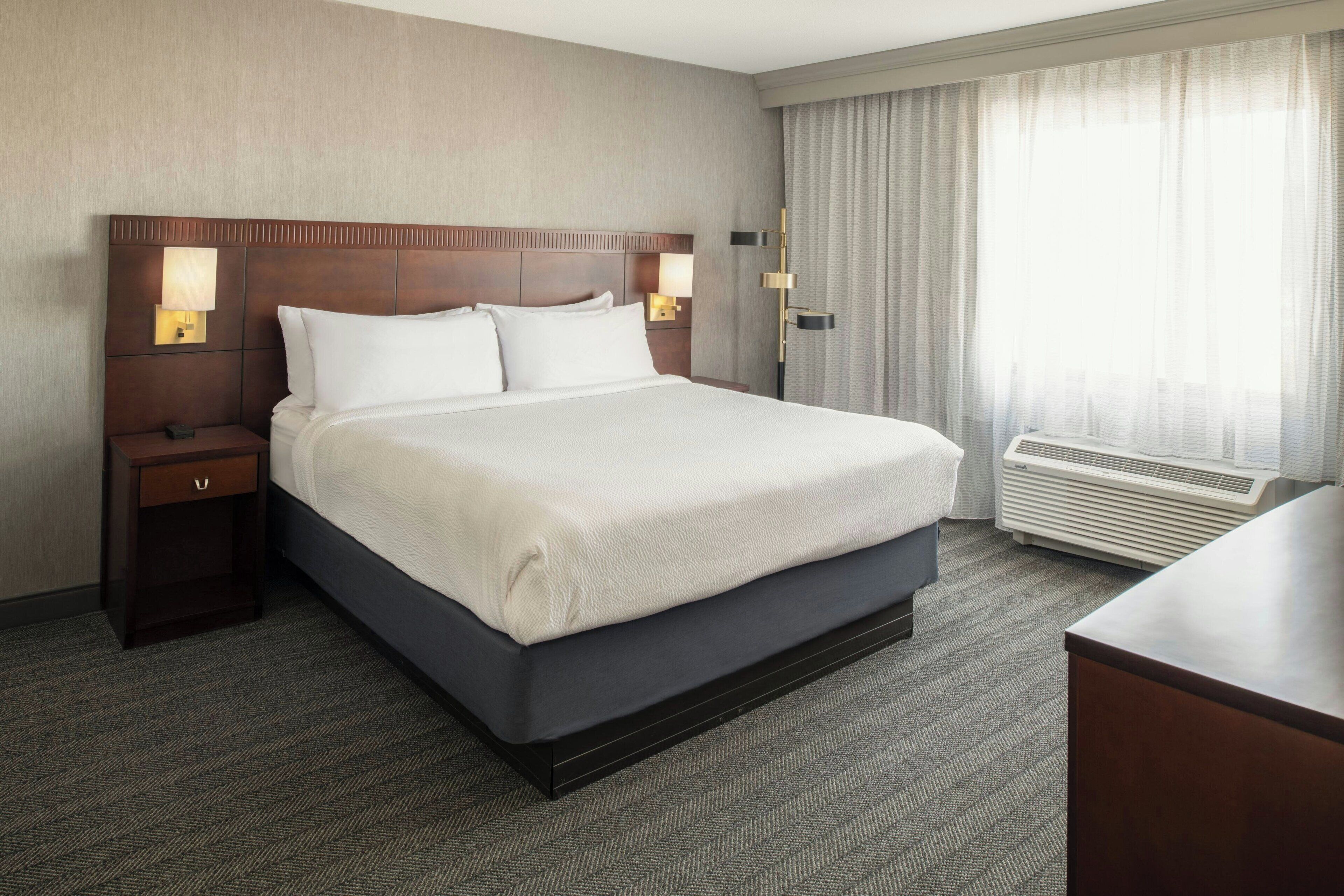 Quarto de hotel com uma cama grande, duas mesas de cabeceira com luminárias, uma escrivaninha e uma janela com cortinas.