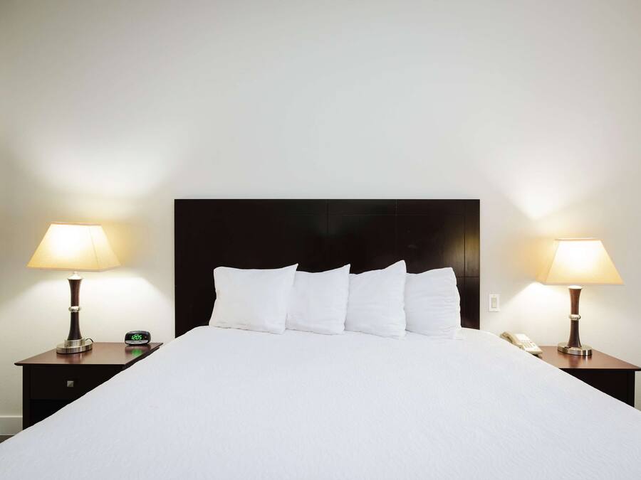 Uma cama bem arrumada com lençóis brancos, duas mesas de cabeceira com abajures e um relógio digital em uma delas.