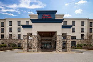 Entrada de um hotel com fachada de pedra, esquema de cores azul e branco e uma placa com a inscrição 'Hampton Inn & Suites'.