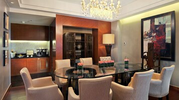 Uma sala de jantar moderna com uma mesa de vidro rodeada por cadeiras acolchoadas, um candelabro e uma cozinha ao fundo.