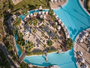 Vista aérea de um resort com piscina central, cercada por palmeiras, espreguiçadeiras e guarda-sóis.