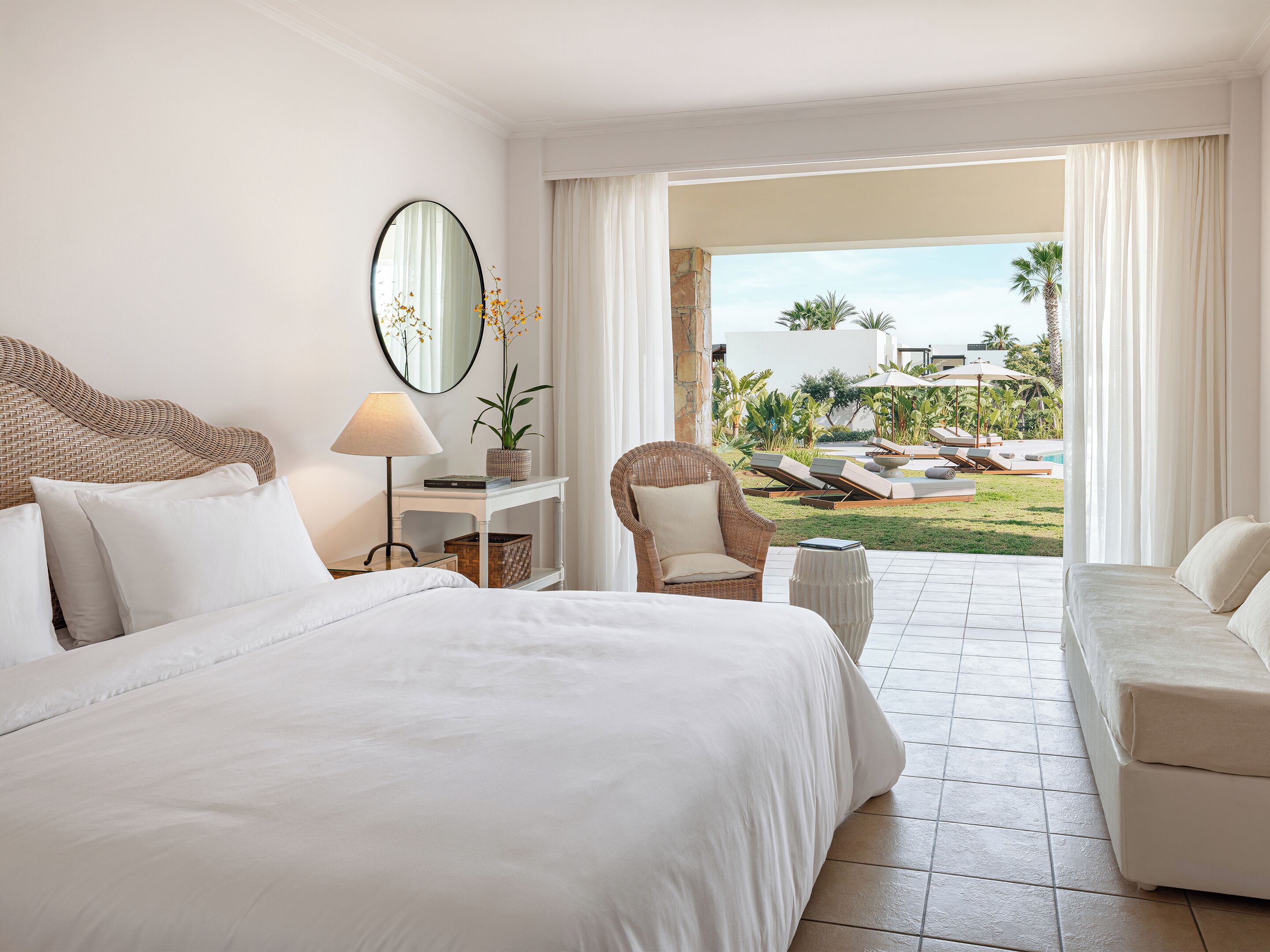 Quarto de hotel com uma cama grande, uma mesa, uma cadeira e vista para uma piscina e jardim.