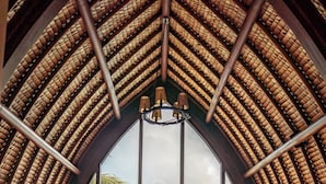 Quarto com teto alto e abobadado feito de materiais trançados, grandes janelas com vista para uma praia tropical e águas azul-claras.