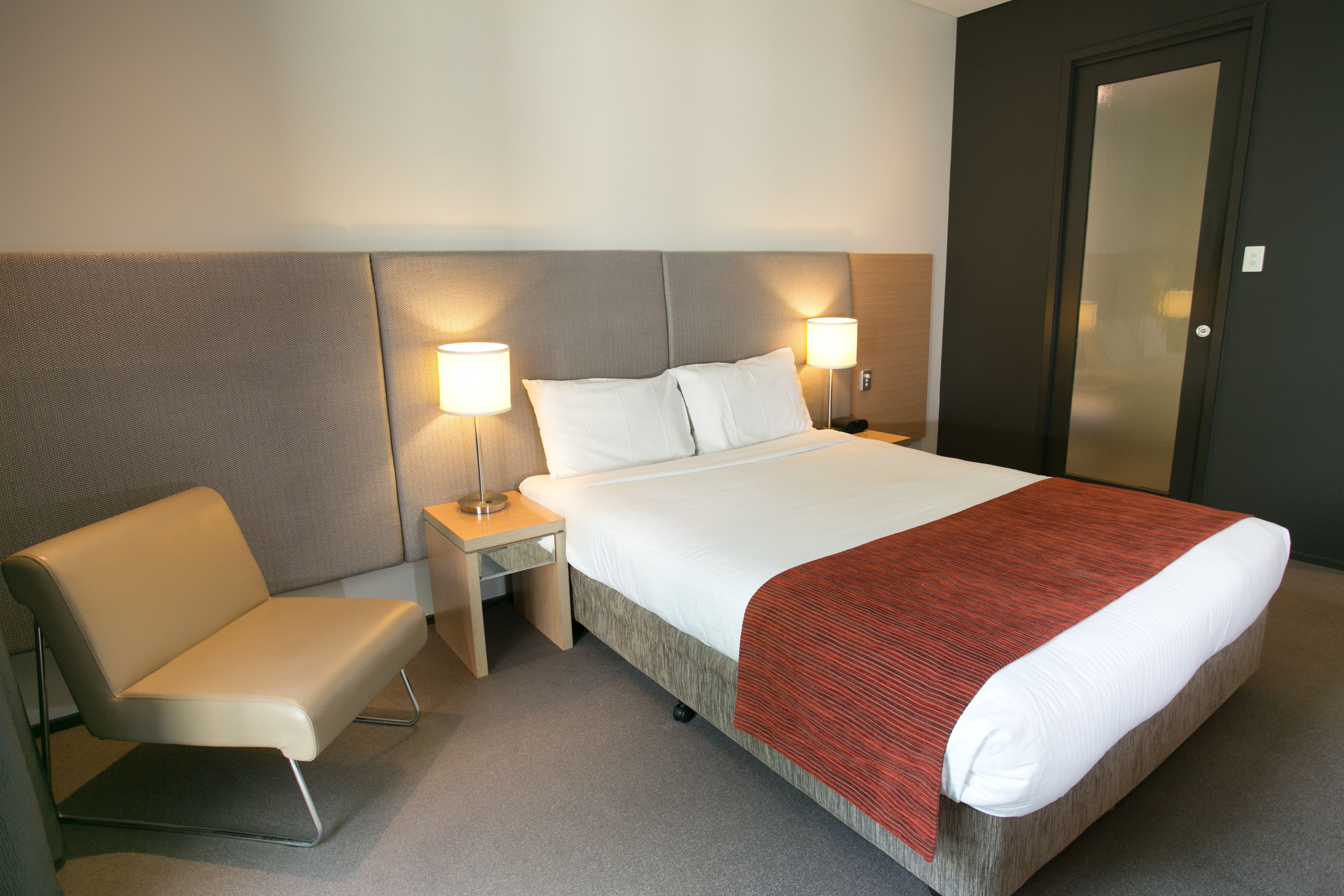 Quarto de hotel com cama, mesa de cabeceira, cadeira e duas luminárias.