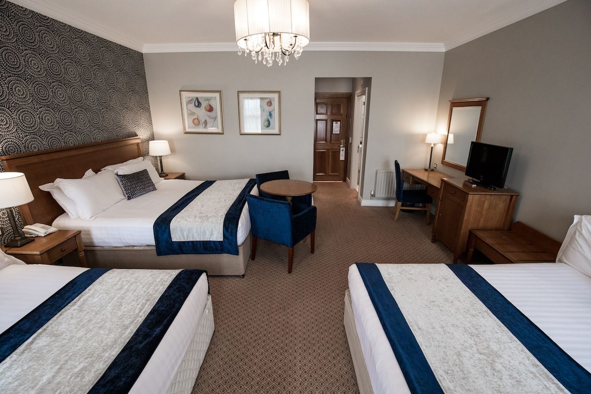 Quarto de hotel com duas camas, uma escrivaninha, uma cadeira, uma televisão e um candelabro.