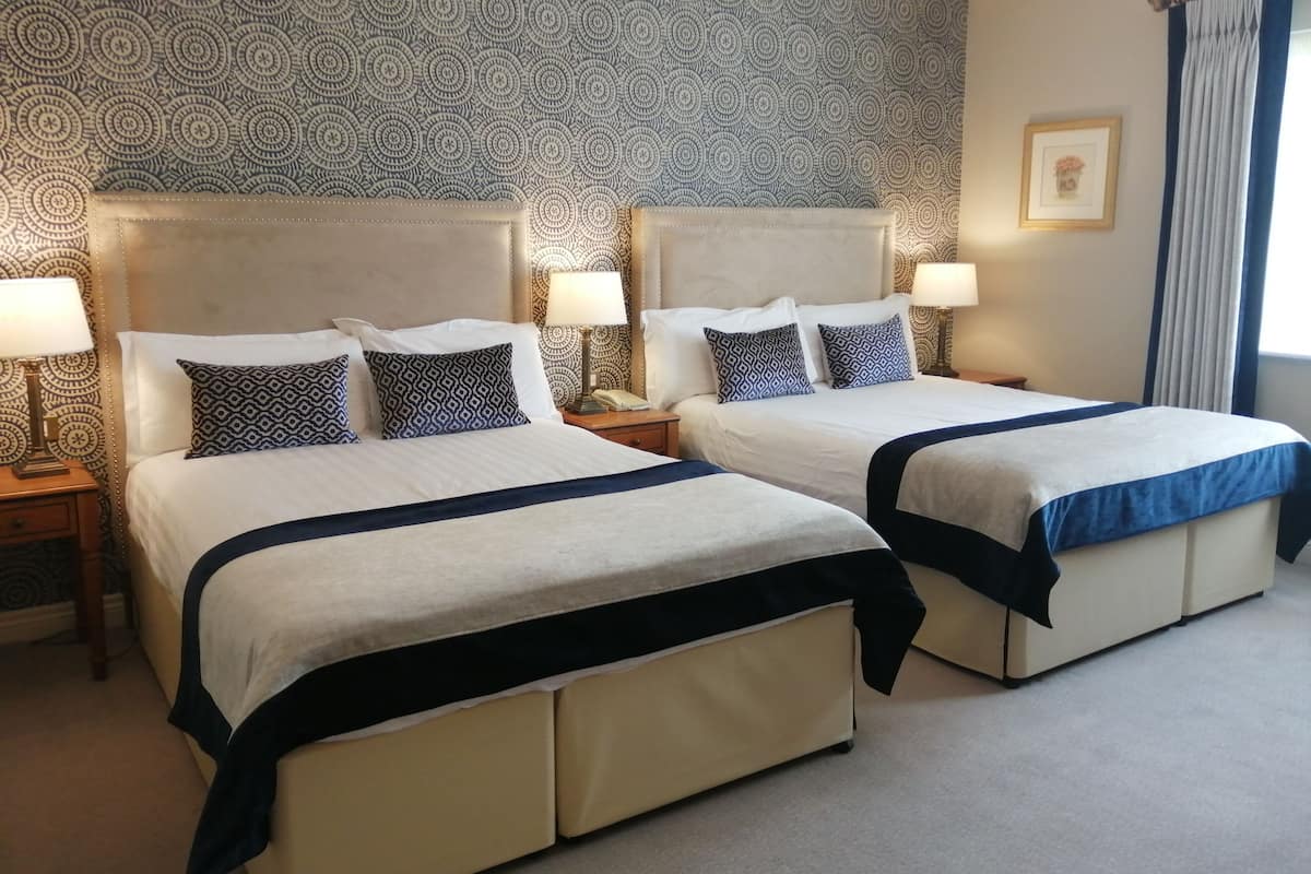 Quarto de hotel com duas camas, cada uma com cabeceira, mesas de cabeceira, luminárias e papel de parede estampado.