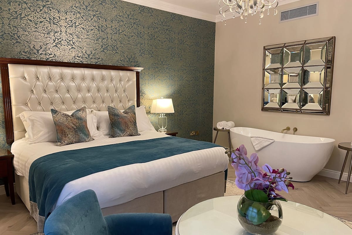 Quarto com cabeceira estofada, cama grande com colcha azul, mesa redonda com um vaso de flores, banheira e espelho com molduras geométricas.