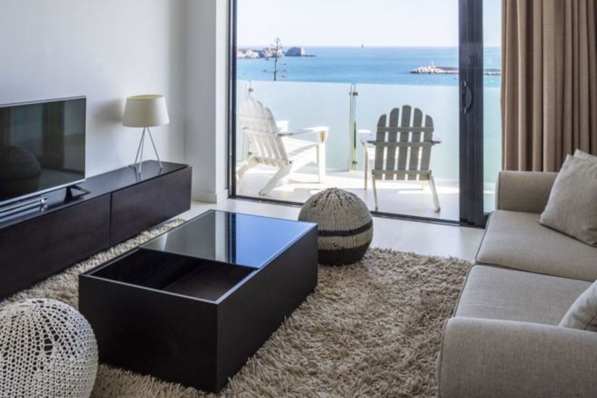 Uma sala de estar moderna com uma televisão grande, uma mesa de centro de vidro e vista para o oceano através de amplas janelas.