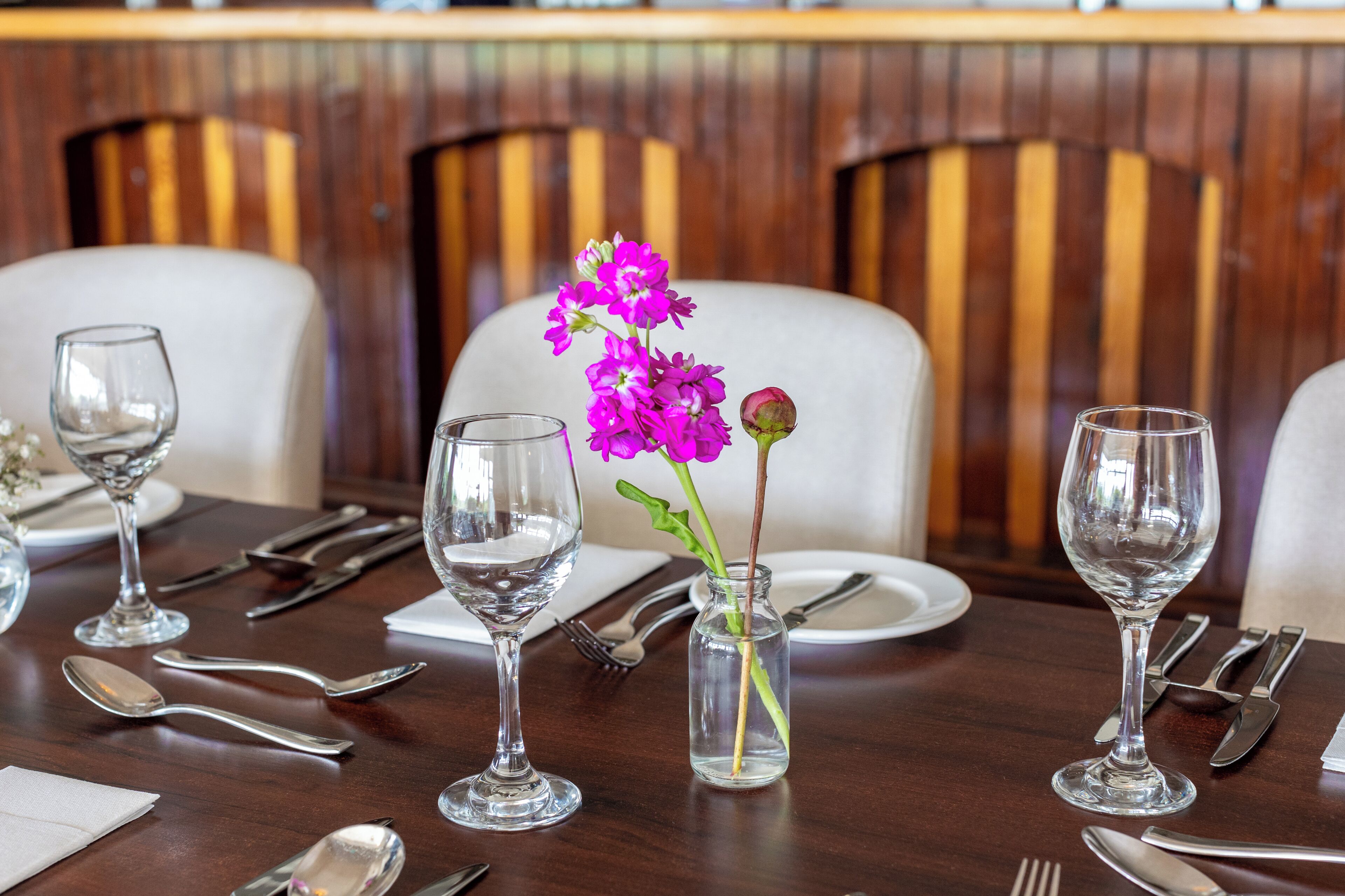 Mesa de jantar posta com copos de vidro transparente, um pequeno vaso com uma flor roxa e talheres bem organizados.