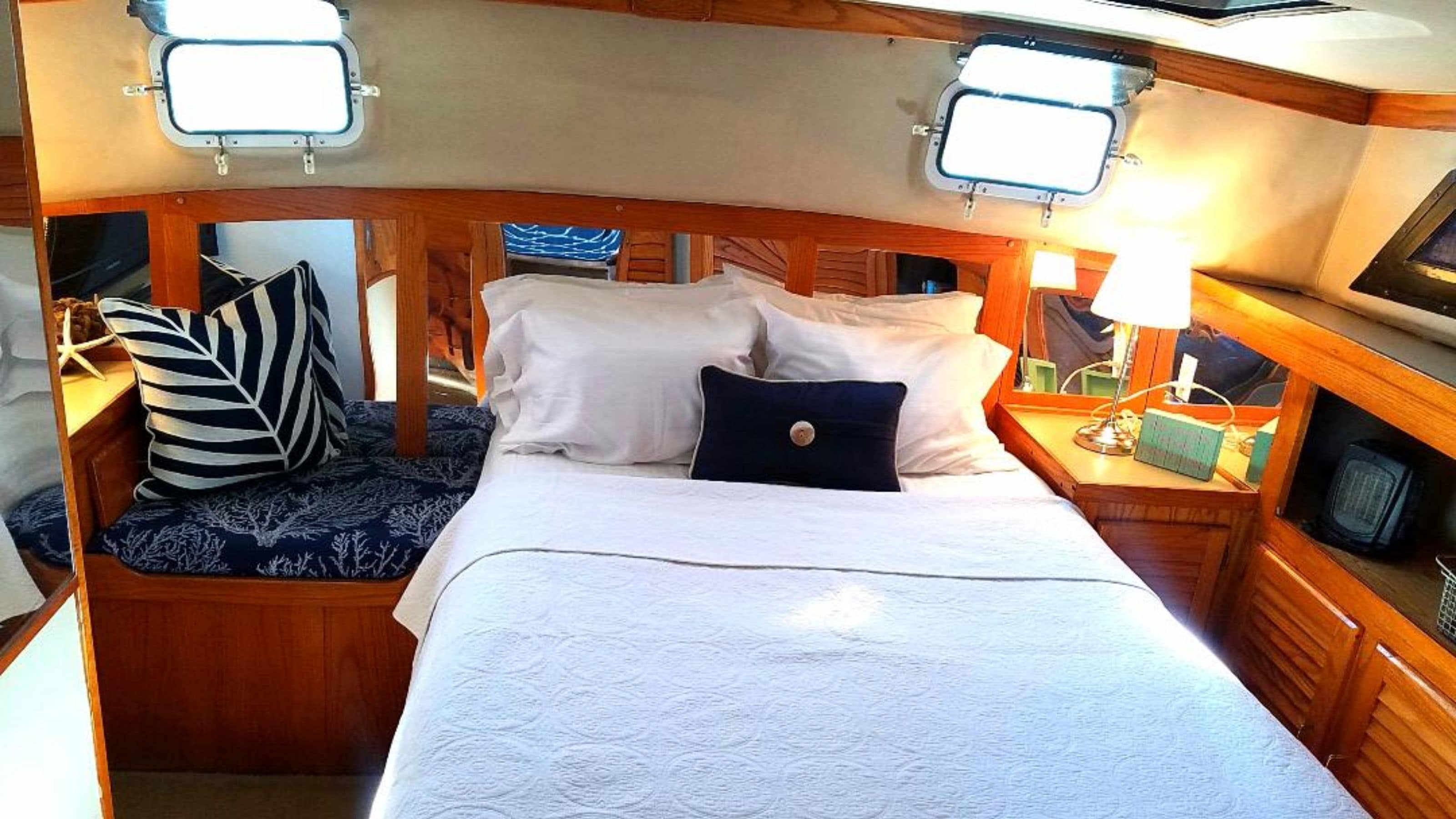 Um quarto compacto e bem iluminado, com uma cama, travesseiros, uma mesinha com abajur e uma televisão.