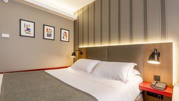 Quarto de hotel com uma cama grande, uma mesa de cabeceira com abajur e um telefone. Há três quadros na parede.