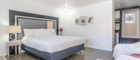 Quarto de hotel moderno com uma cama grande, mesa de cabeceira com abajur e um pequeno espaço de estar.