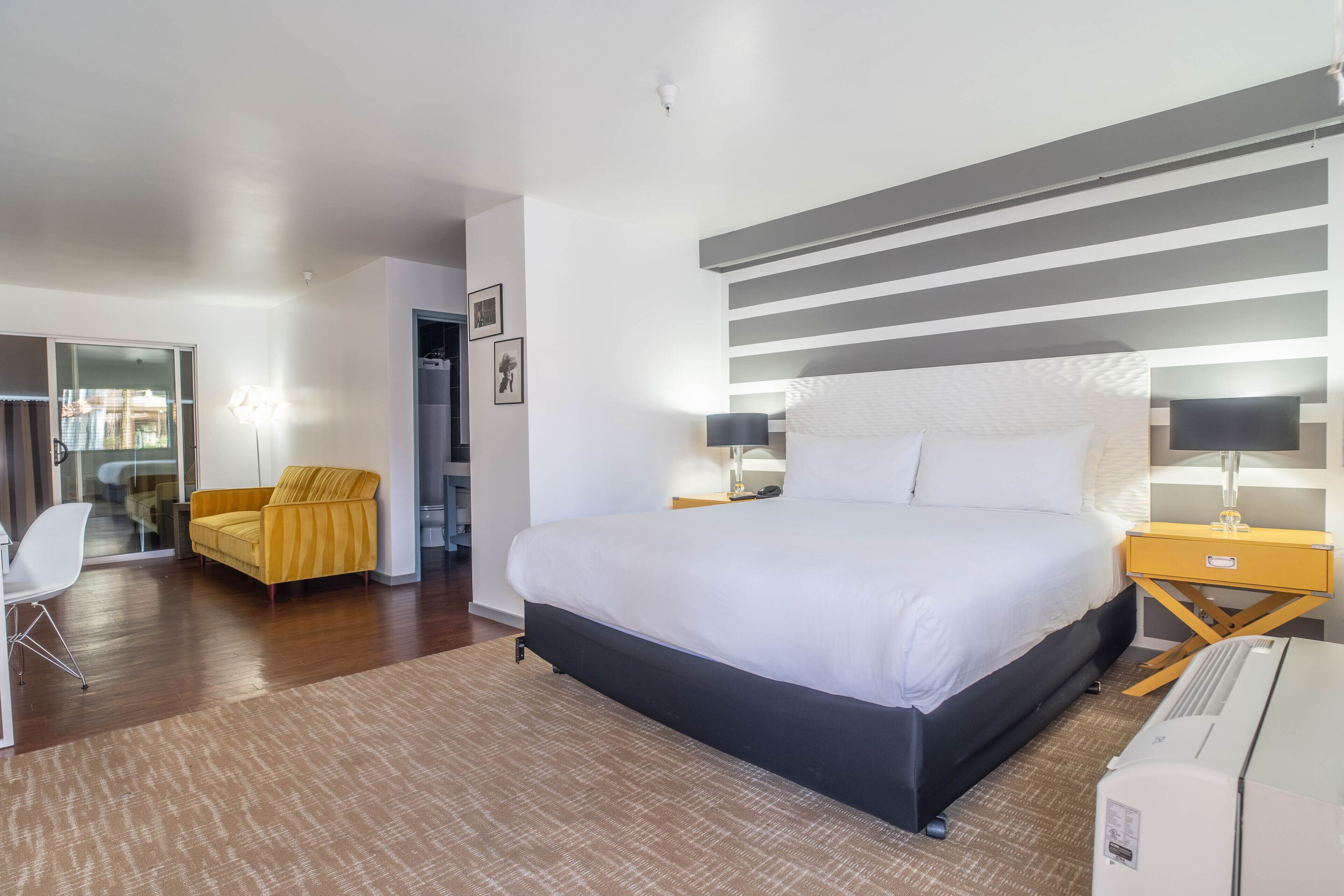 Quarto de hotel moderno com uma cama grande, uma poltrona amarela, uma escrivaninha com abajur e vista para a cidade através de uma porta de vidro.