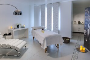 Um salão de spa moderno com mesa de massagem, maca de tratamento, escrivaninha com velas e uma grande janela com cortinas.