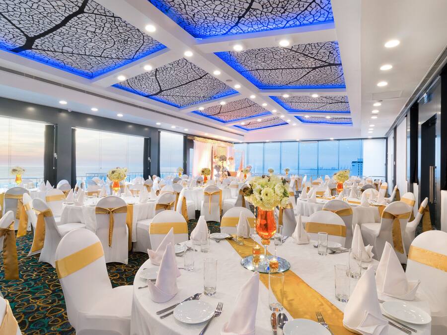 Salão de banquetes com mesas redondas preparadas para um evento formal, com toalhas brancas, fitas douradas e arranjos florais.