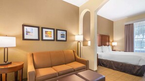 Quarto de hotel com sofá, mesa pequena, cama com cabeceira e quadros na parede.