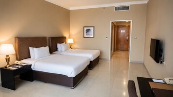Quarto de hotel com duas camas, uma escrivaninha, uma cadeira, uma televisão e uma porta que dá para outro cômodo.