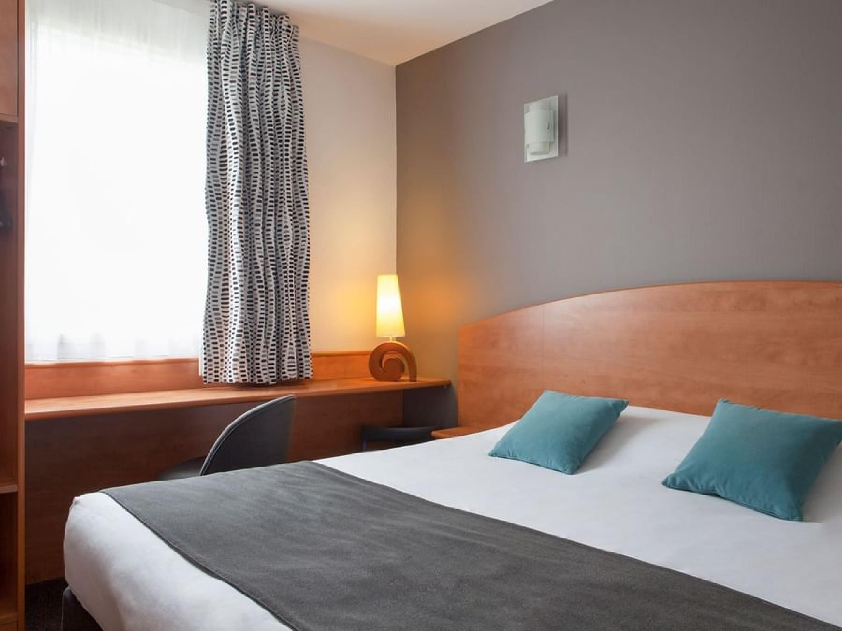Quarto de hotel com cama, escrivaninha, cadeira, luminária e janela com cortinas.