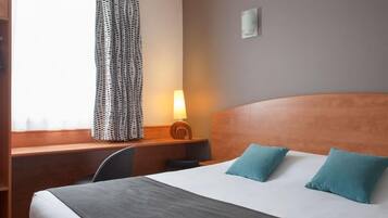 Quarto de hotel com cama, escrivaninha, cadeira, luminária e janela com cortinas.