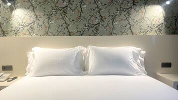 Cama bem arrumada com lençóis brancos, duas mesas de cabeceira com telefone e controle remoto, e um padrão de papel de parede floral na parede.