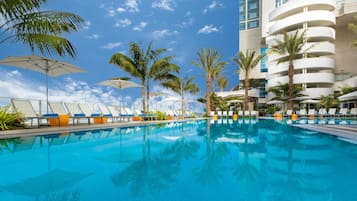 Piscina com espreguiçadeiras e guarda-sóis, cercada por palmeiras e um edifício moderno.