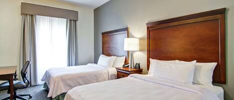 Quarto de hotel com duas camas, uma mesa, uma cadeira, um abajur e uma janela com cortinas.