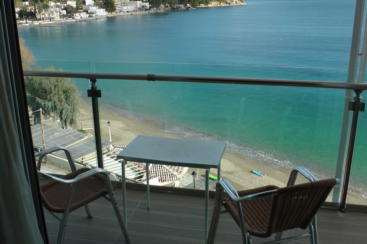 Varanda com mesa e duas cadeiras, vista para uma praia e mar azul-turquesa.