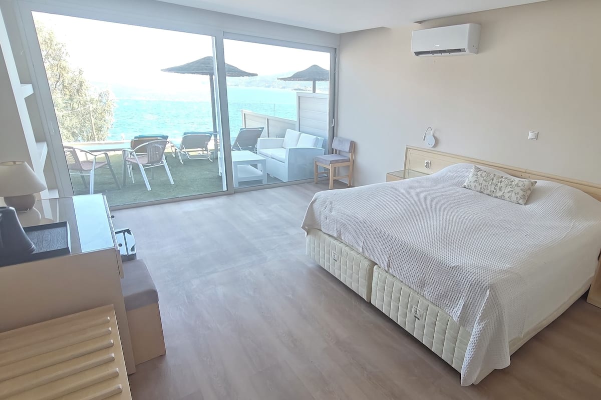 Quarto de hotel com uma cama grande, uma escrivaninha, uma cadeira, uma mesinha e vista para o mar através de uma porta de correr.