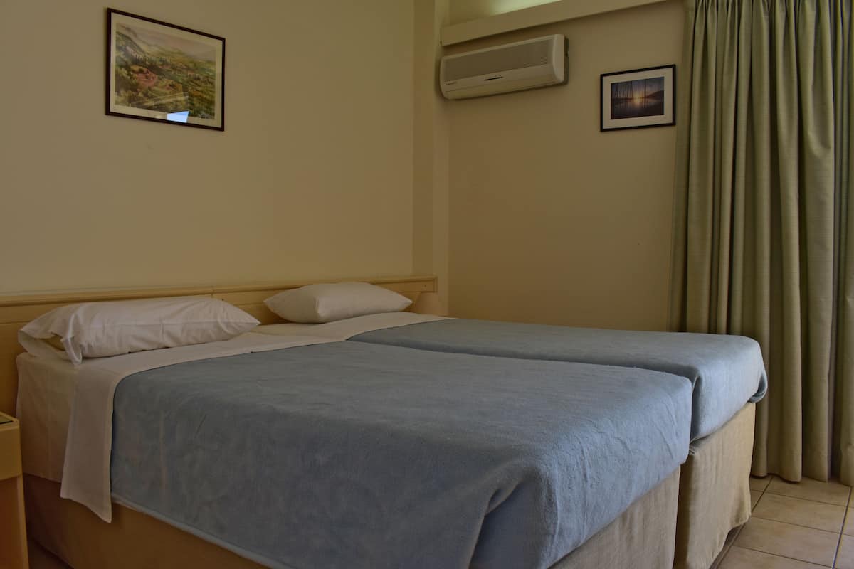Quarto de hotel com duas camas, mesa de cabeceira, aparelho de ar condicionado e janela com cortinas.