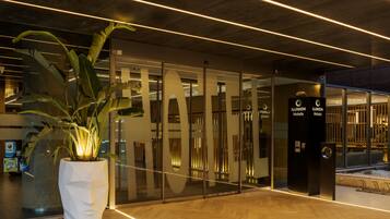 Uma entrada de hotel moderna com uma grande planta em vaso, portas de vidro e sinalização iluminada.