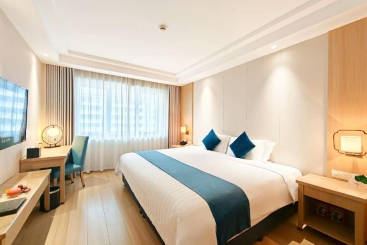 Quarto de hotel com uma cama grande, uma escrivaninha, uma cadeira, uma TV e uma janela com cortinas.